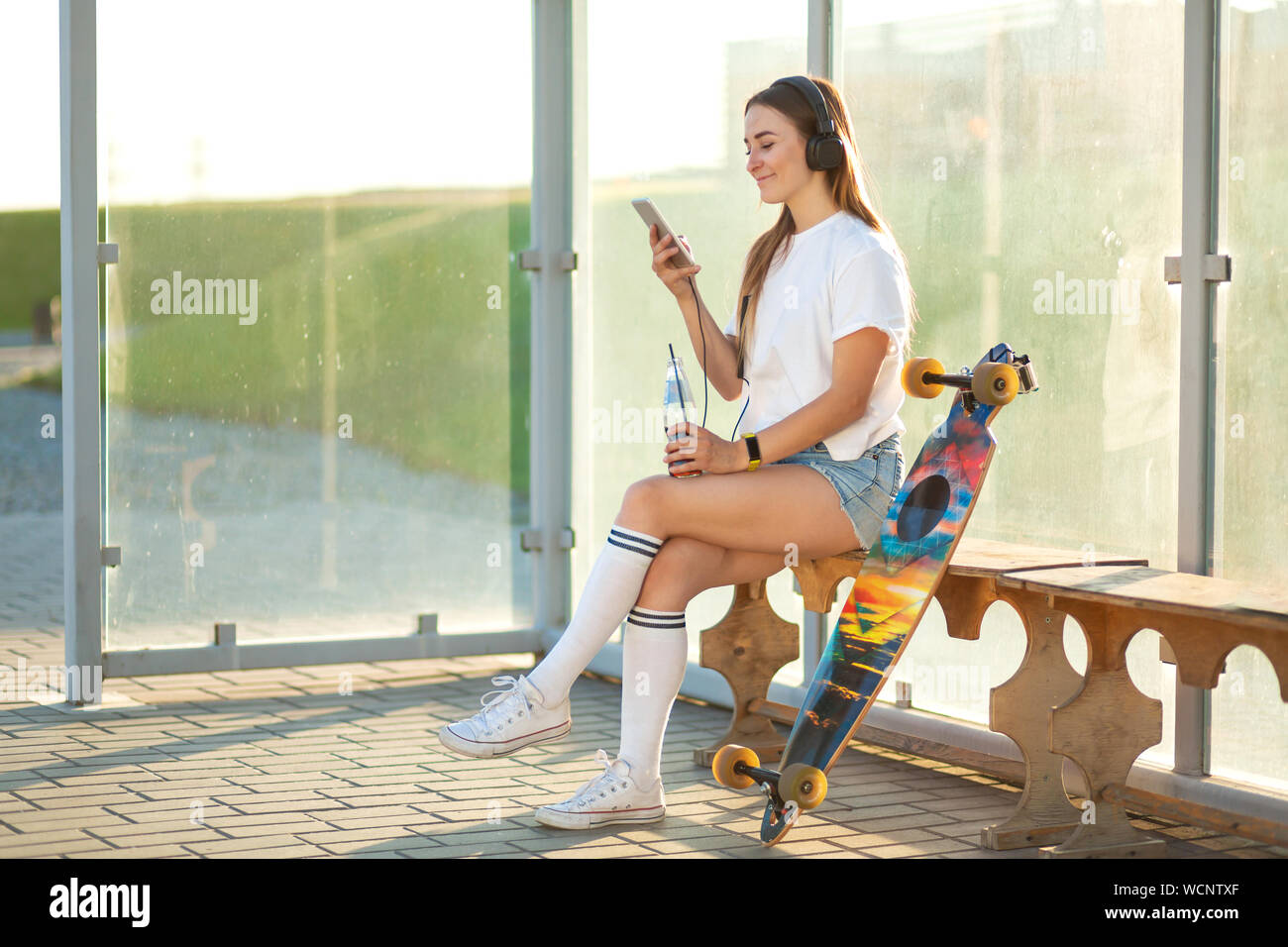 Stylish young girl sitting on bus stop with her longboard, listening ...