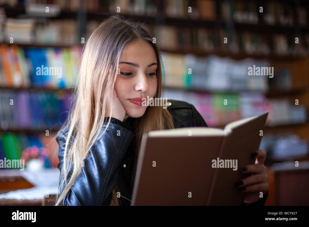 Beautiful teenager schoolgirl sitting in the library and reading a book ...