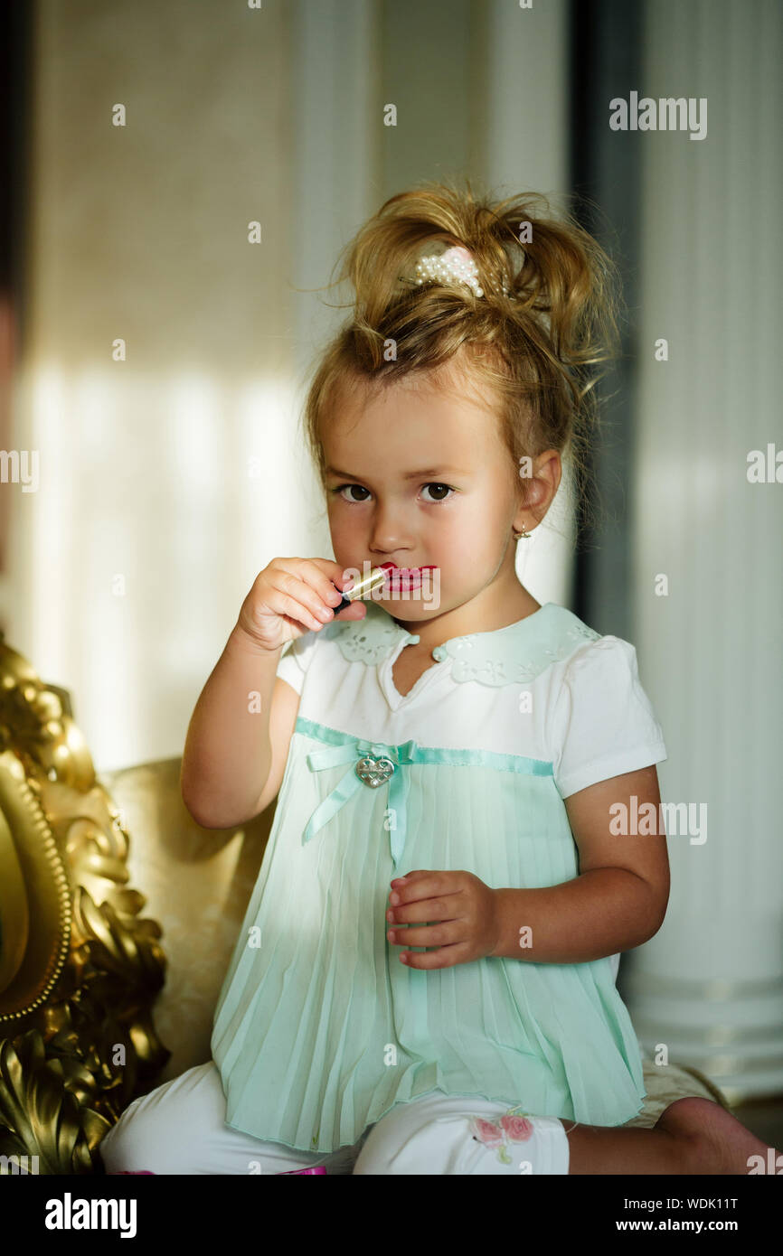 Baby girl put red lipstick on lips. Kid model make up at mirror in room ...