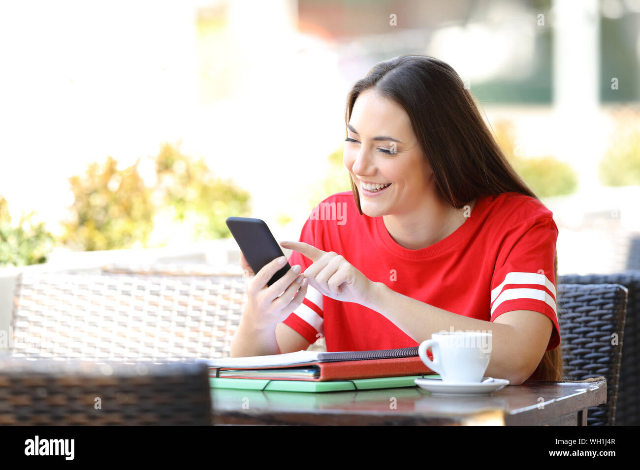 Happy student in red browsing phone content sitting in a coffee shop ...