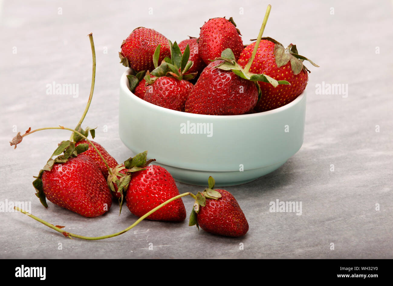 Some strawberries in a light green bowl and some over the surface ...