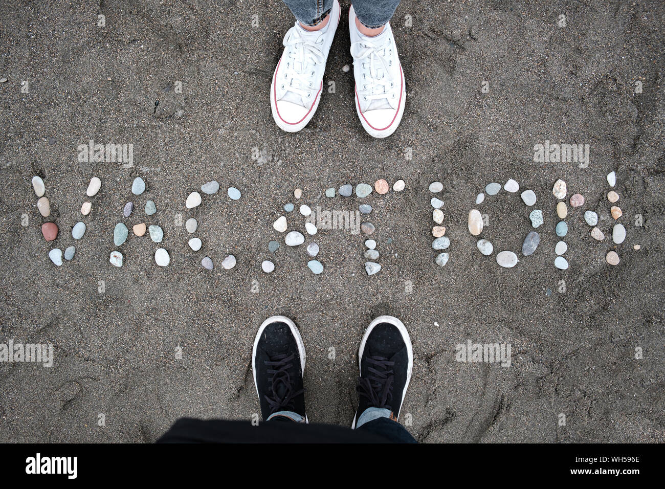Word "vacation" laid out with sea pebble on the beach. Two persons ...