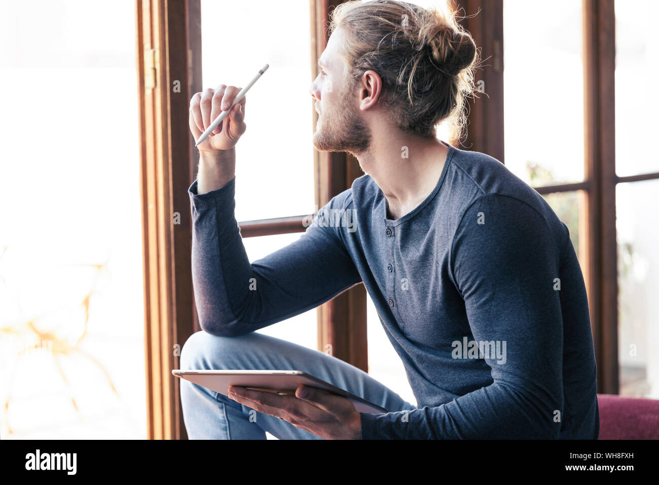 Pensive young man at home with a digital tablet looking out of a window ...