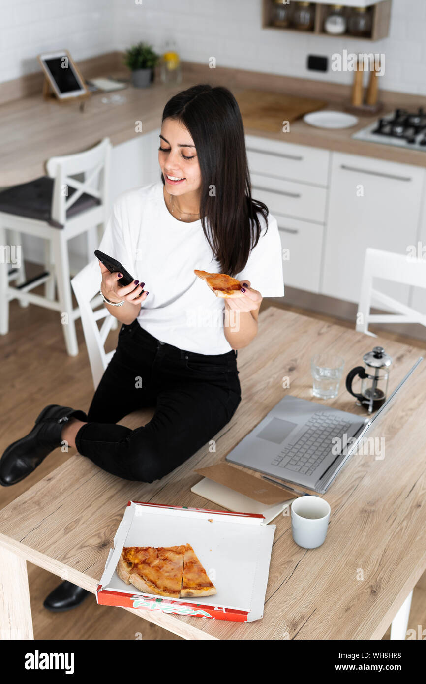 Young woman with cell phone eating pizza in kitchen at home Stock Photo ...