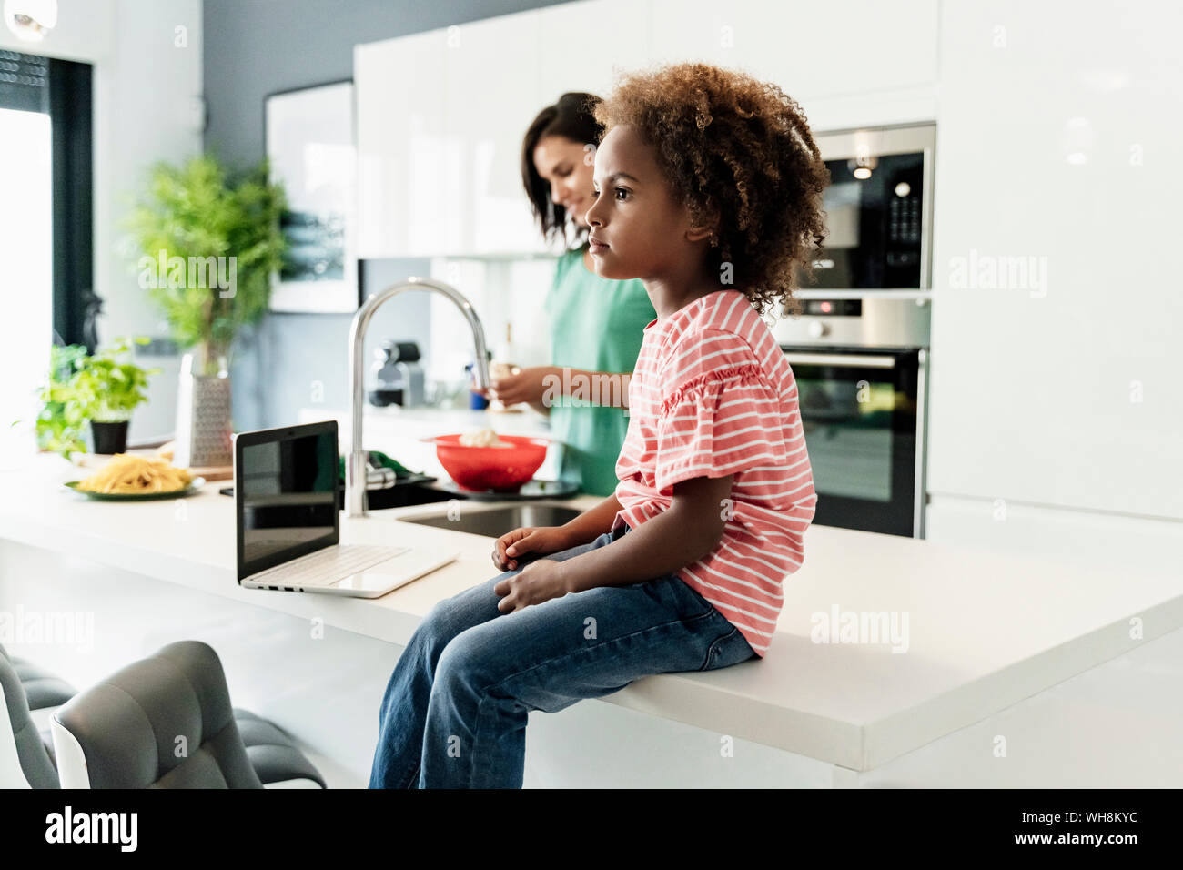 Girl sitting on kitchen counter with laptop and mother in background ...