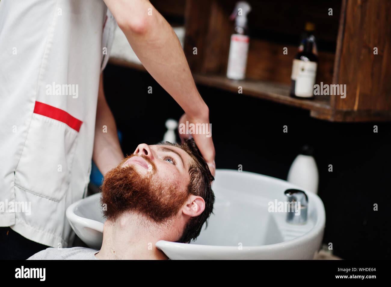 Young bearded man washing head by hairdresser while sitting in chair at ...