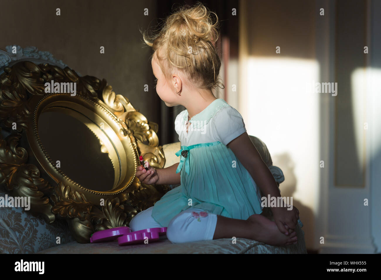 Baby girl put red lipstick on lips. Kid model make up at mirror in room ...