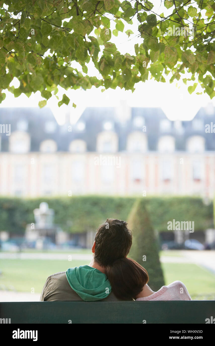 Rear view of a woman leaning head on man's shoulder on bench in park ...