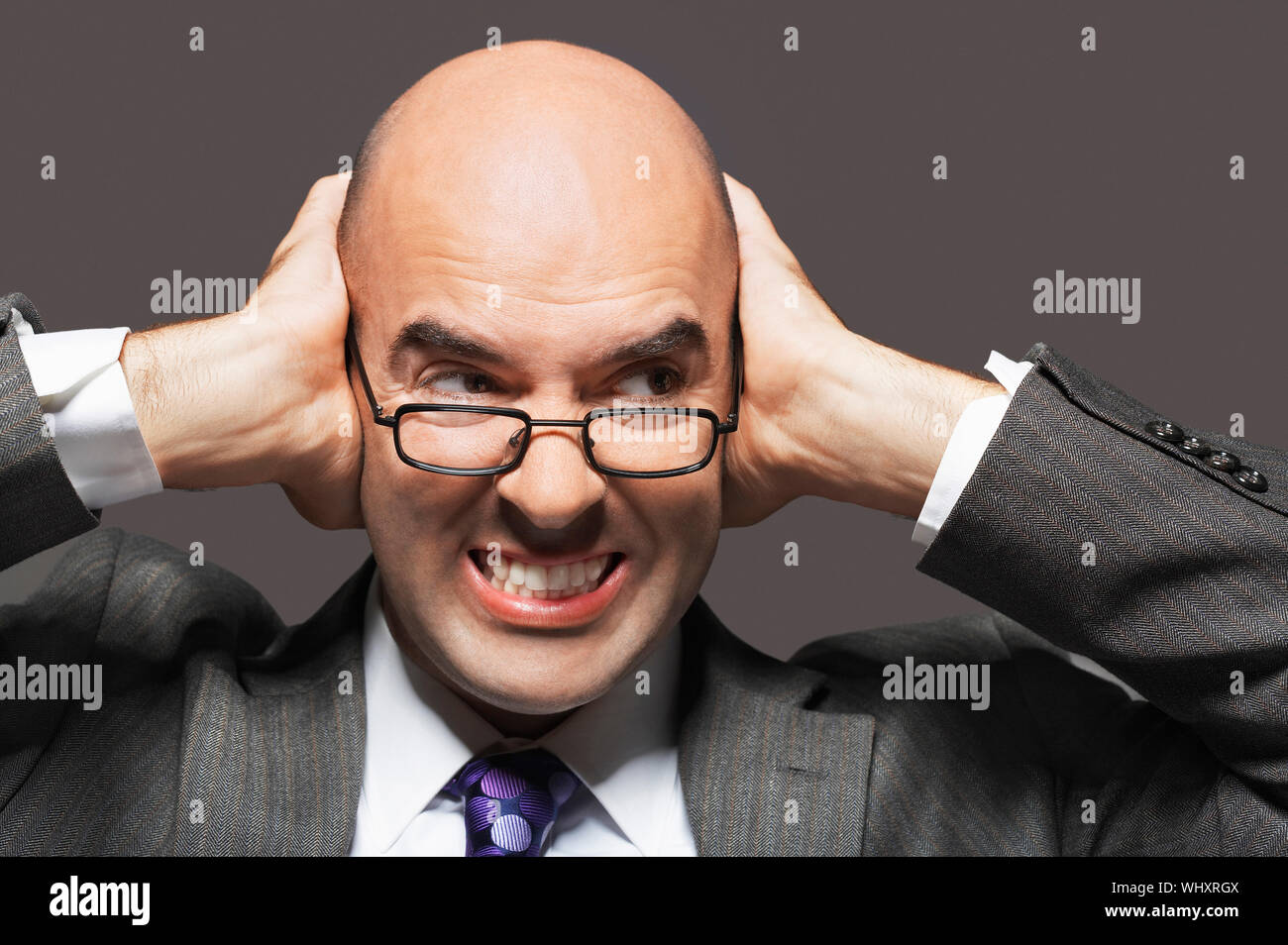 Bald businessman with hands over ears looking sideways against gray ...