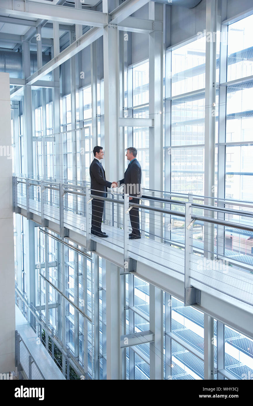 Full length of businessmen shaking hands by railing in modern office ...