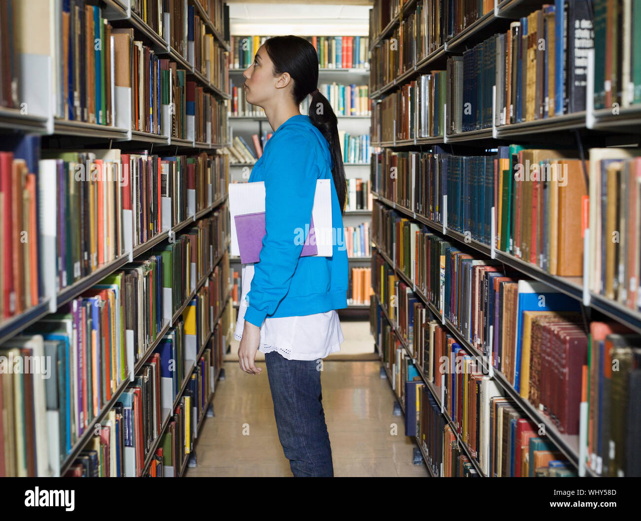 Side view of a young woman standing by bookshelf in library Stock Photo ...