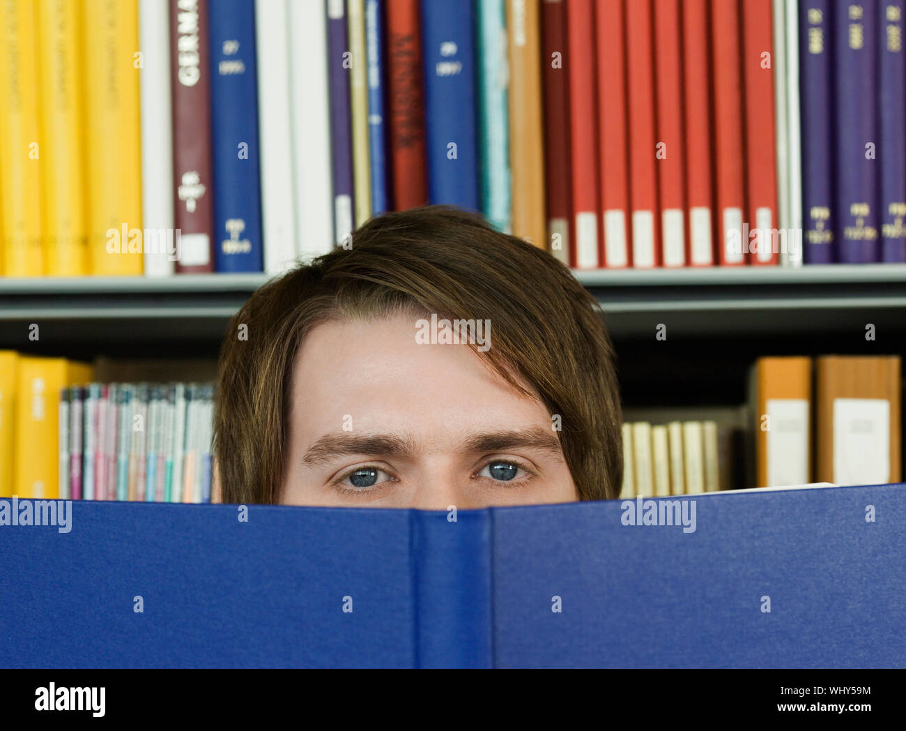 Closeup portrait of a young man peeking over opened book in library ...