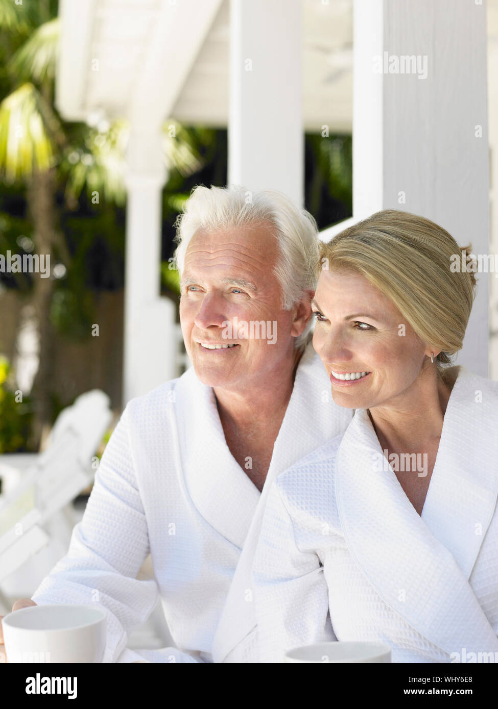 Middle aged couple in bathrobes with coffee cups sitting on verandah ...