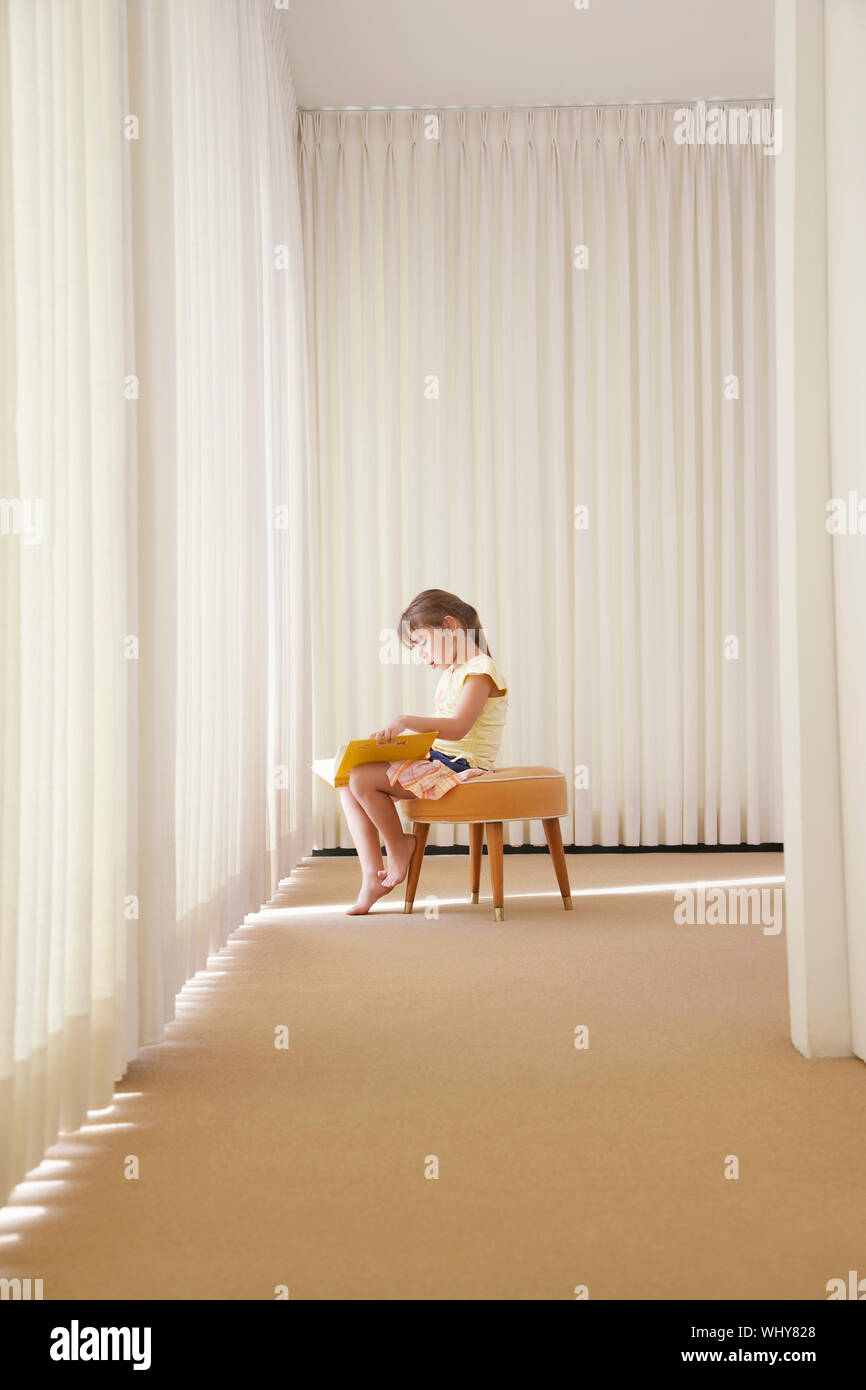 Side view of little girl reading book on stool in corner of room Stock ...