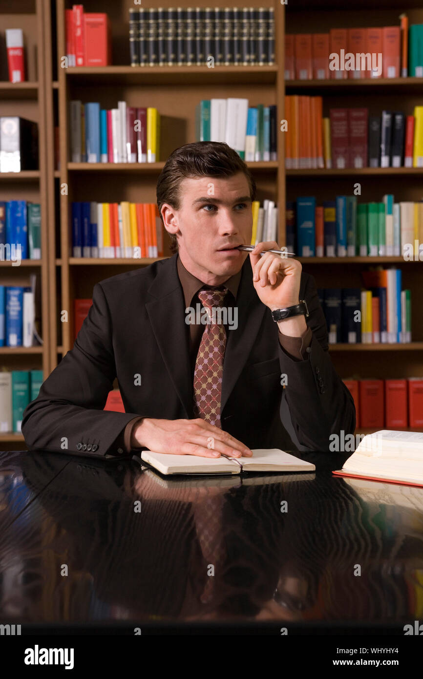 Thoughtful young man in formals chewing pen at desk in library Stock ...