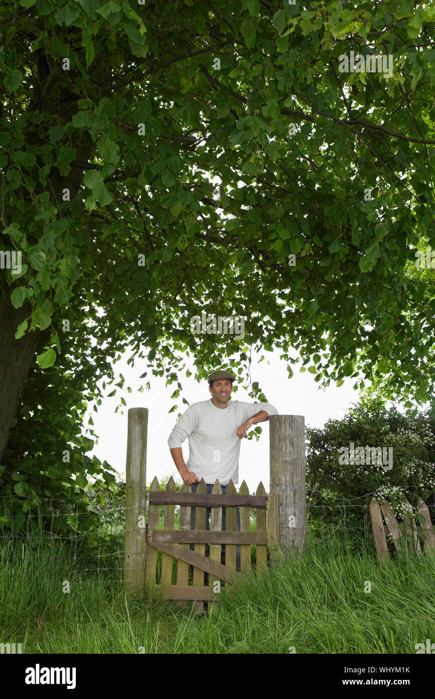 Portrait of a smiling man standing across field gate under the tree ...
