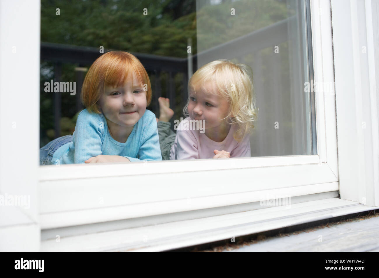 Portrait of cute girl lying with sister in front of glass window at ...
