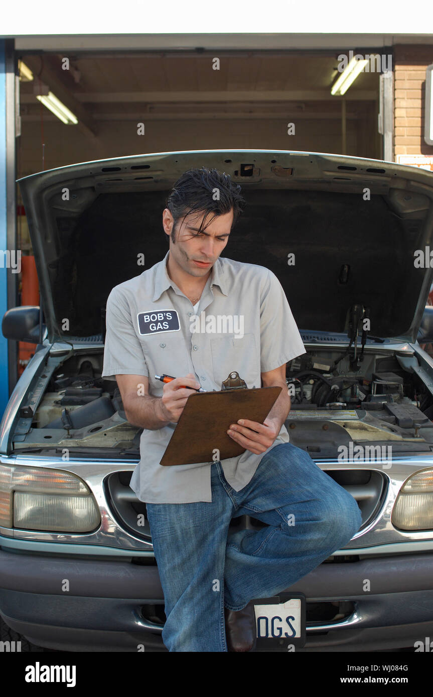 Mechanic writing on clipboard by open car hood Stock Photo - Alamy