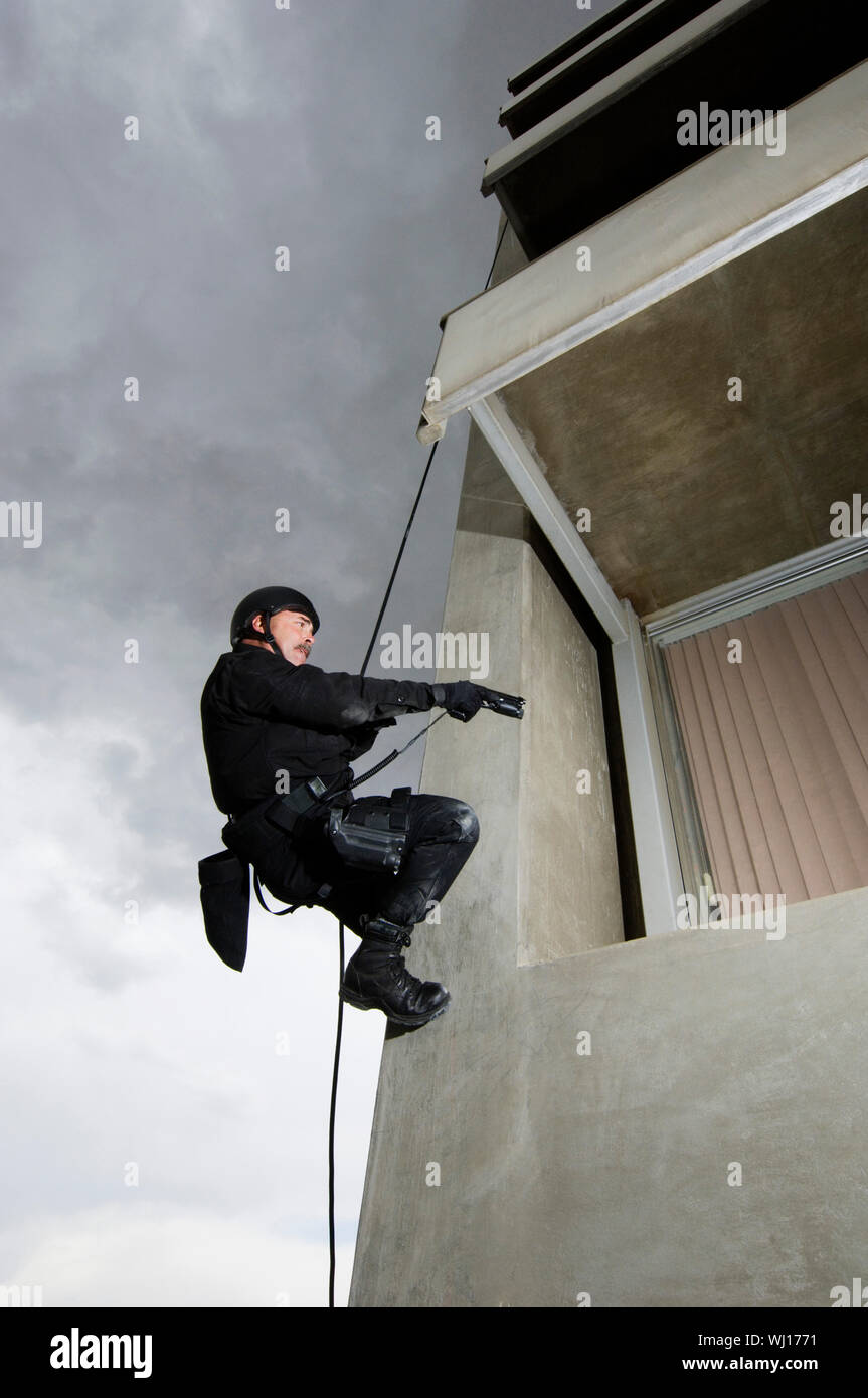 Full length of a trained man aiming gun while rappelling down the rope ...