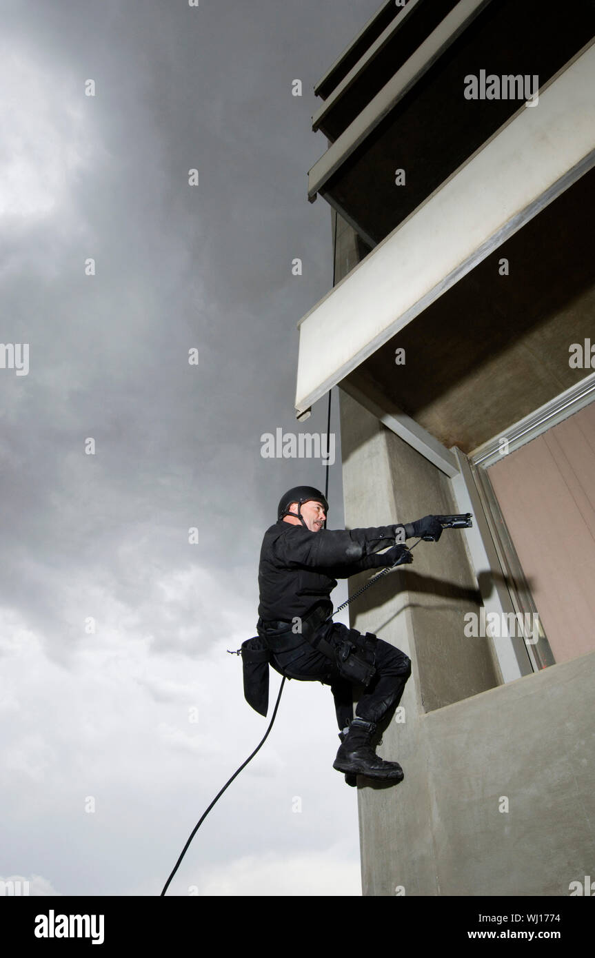 Full length of a trained man aiming gun while rappelling down the rope ...
