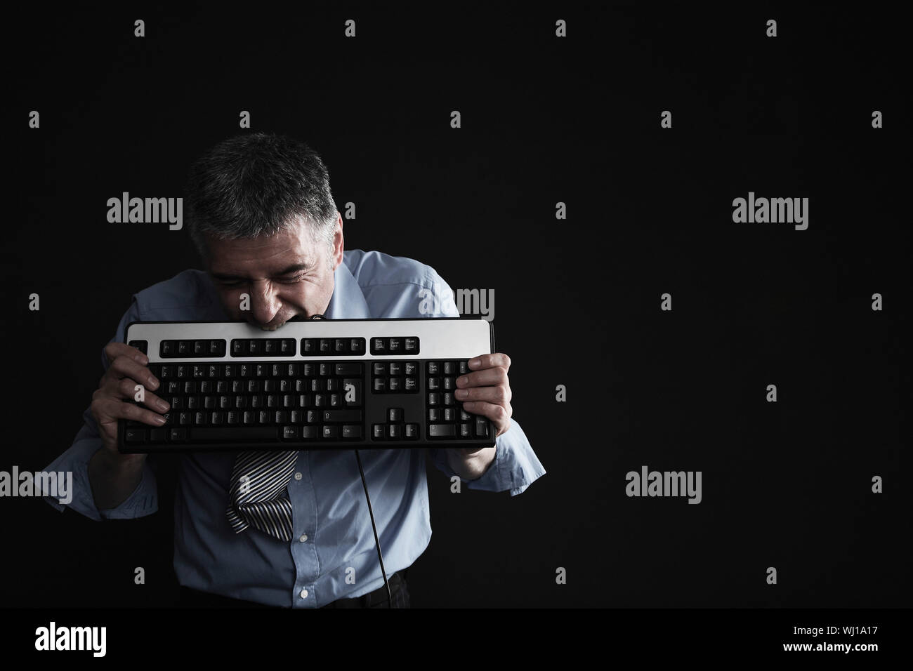 Worried middle aged businessman biting computer keyboard against black ...