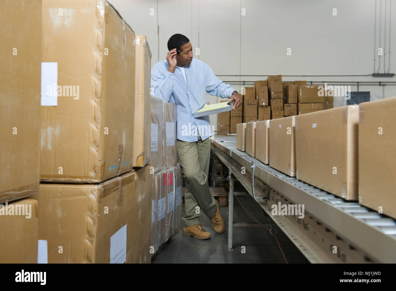 Full length of a man standing by conveyor belt and boxes with clipboard ...
