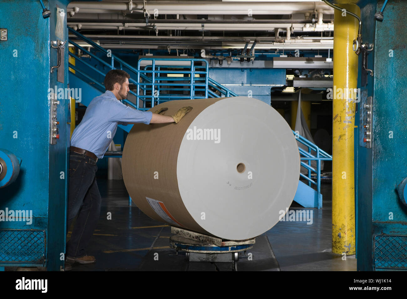 Side view of a man pushing huge roll of paper in newspaper factory ...