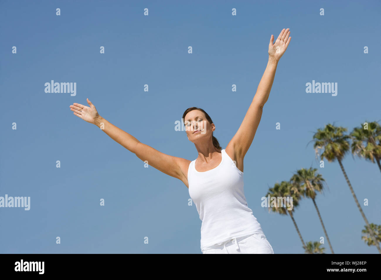 Low angle view of mature woman in white sportswear meditating with arms ...