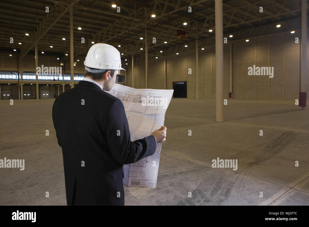 Rear view of male architect looking at blueprint in empty warehouse ...