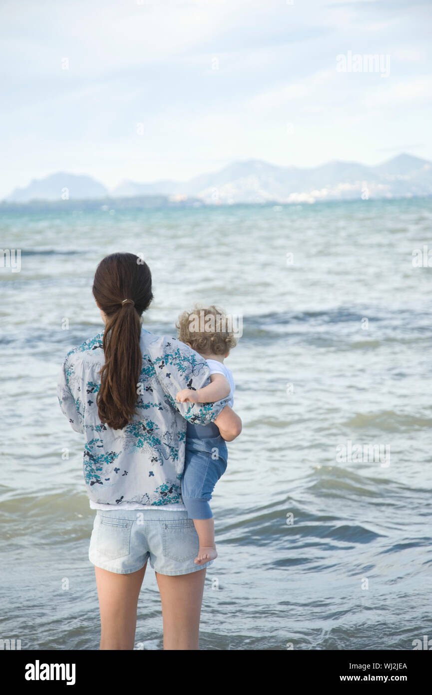 Rear view of mother with baby looking at sea Stock Photo - Alamy