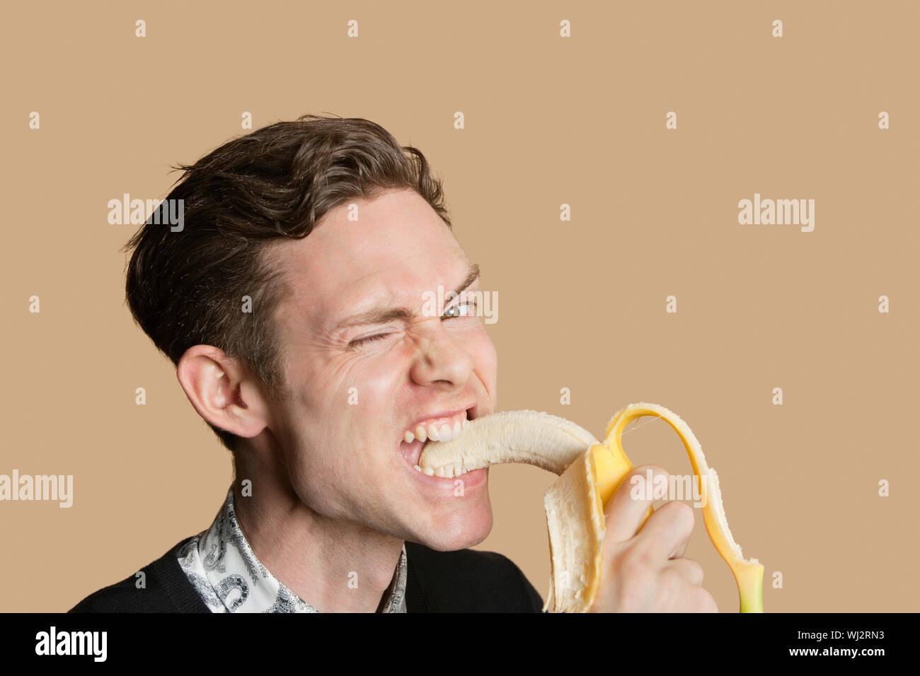 Portrait of a mid adult man winking while biting banana over colored ...