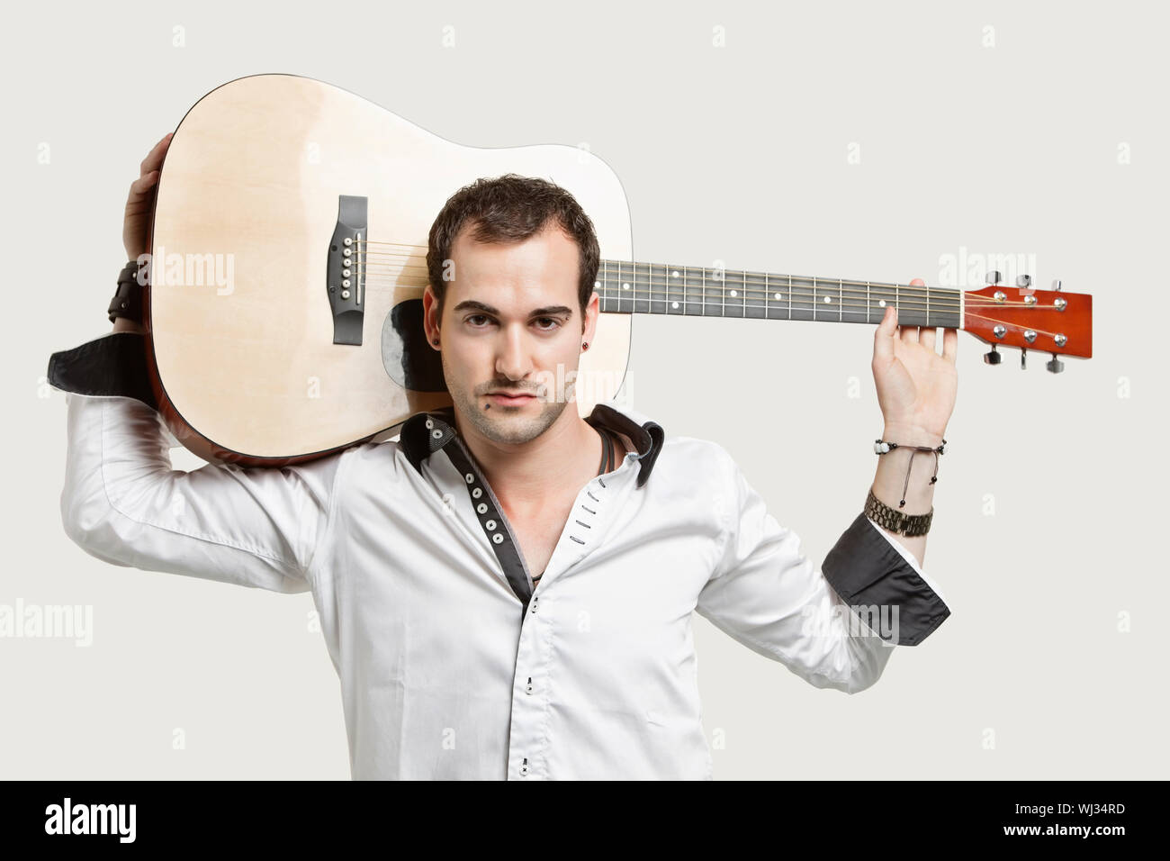 Portrait of young man carrying guitar over his shoulder against gray ...