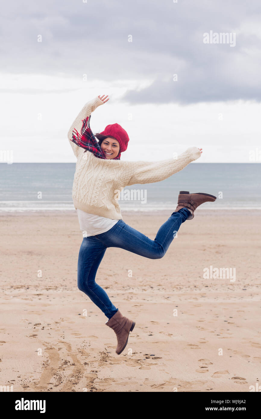 Full length of a woman in stylish warm clothing jumping at the beach ...