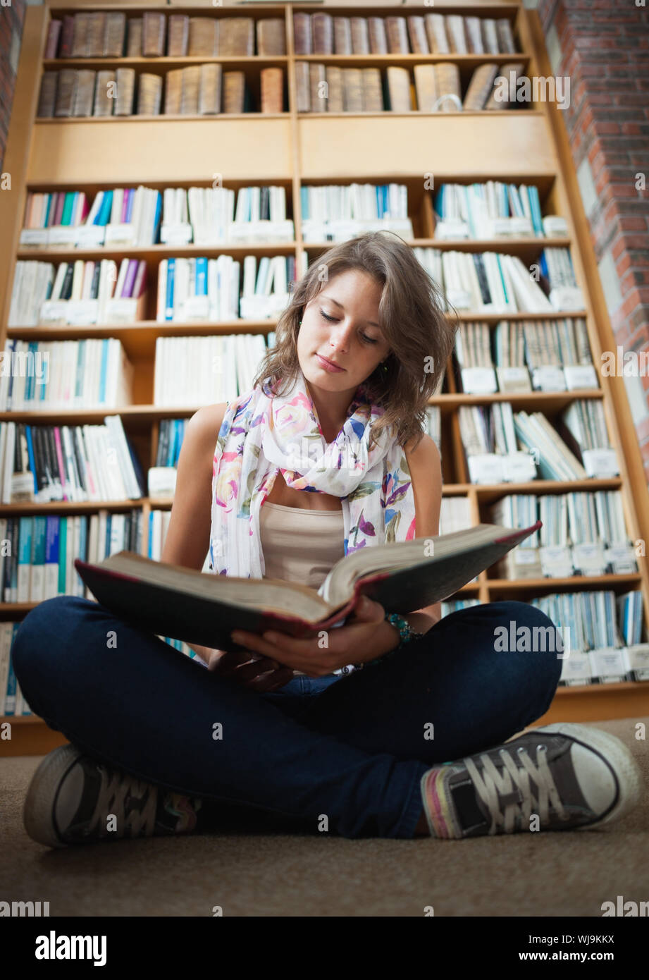 Full length of a female student sitting against bookshelf and reading a ...