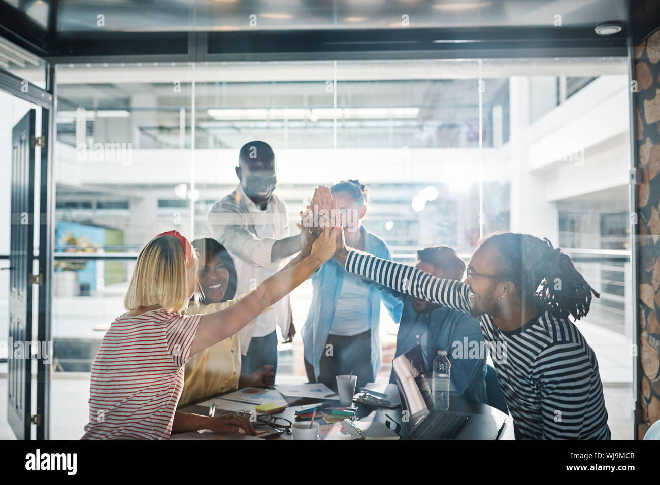 Laughing group of diverse businesspeople high fiving each other during a meeting in a glass ...