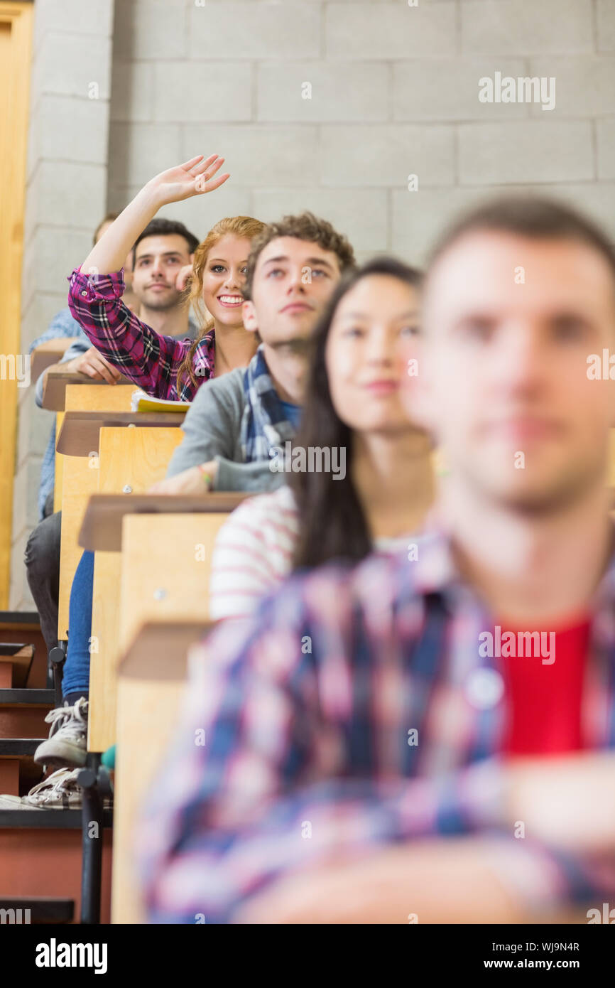Smiling young female student raising hand by others in a row at the ...