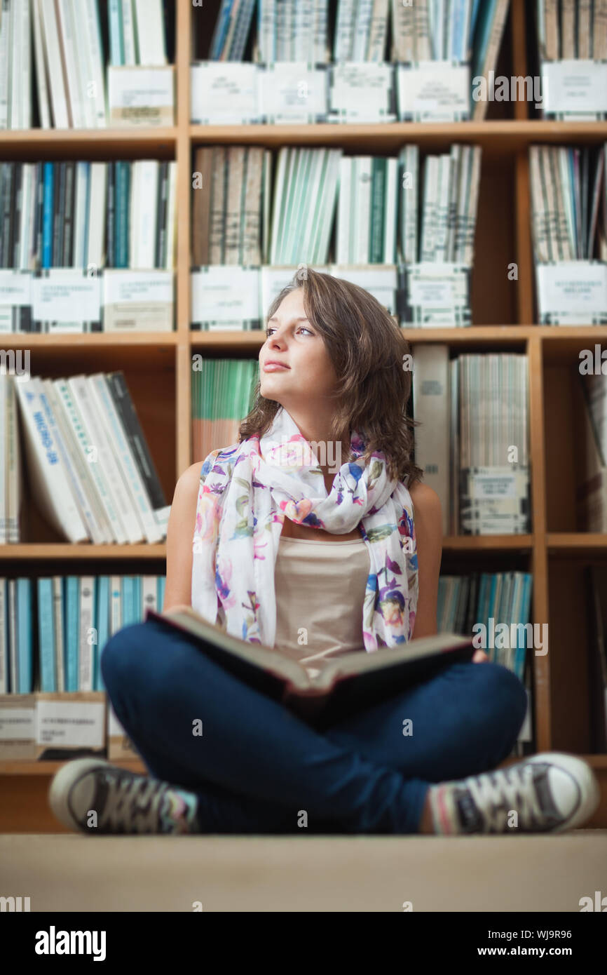 Thoughtful female student sitting against bookshelf with a book on the ...
