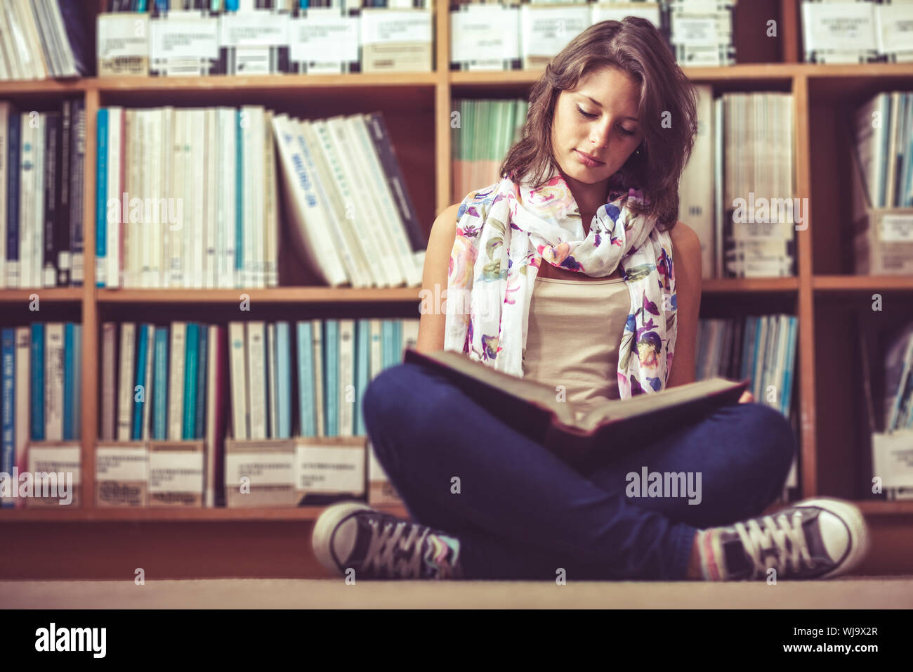 Full length of a female student sitting against bookshelf and reading a ...