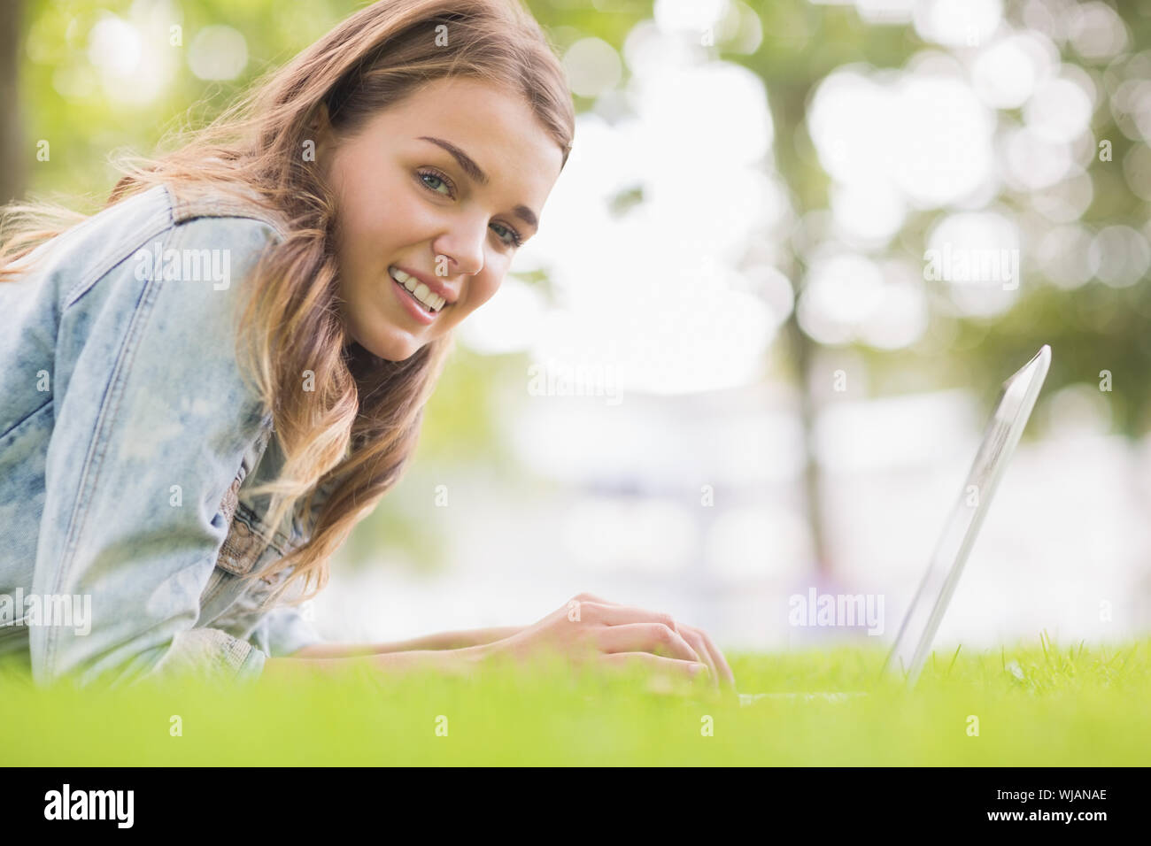 Happy student lying on the grass using her laptop looking at camera ...