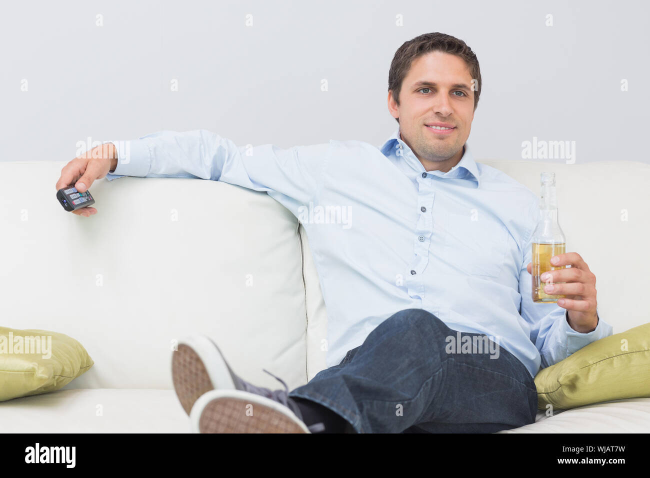 Relaxed man with a drink and remote control sitting on sofa at home ...