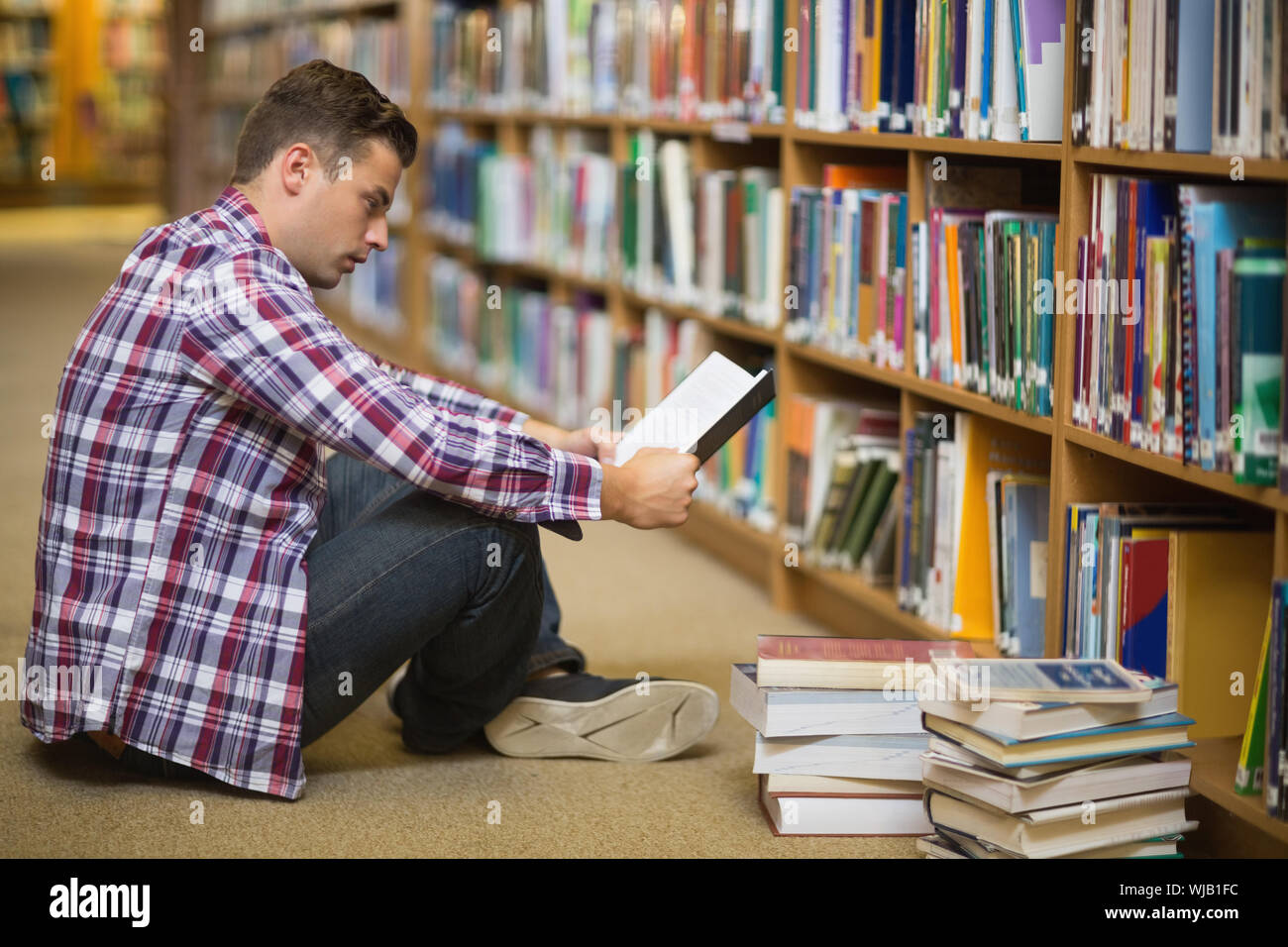 Handsome young student sitting on library floor reading book Stock ...
