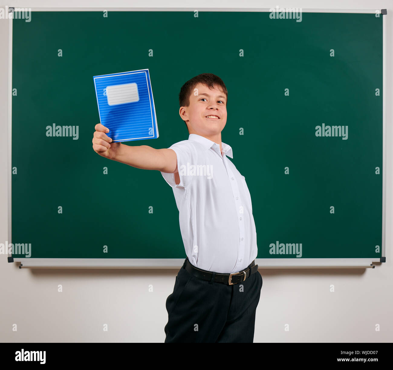 Portrait of a school boy posing with blue exercise book on blackboard ...