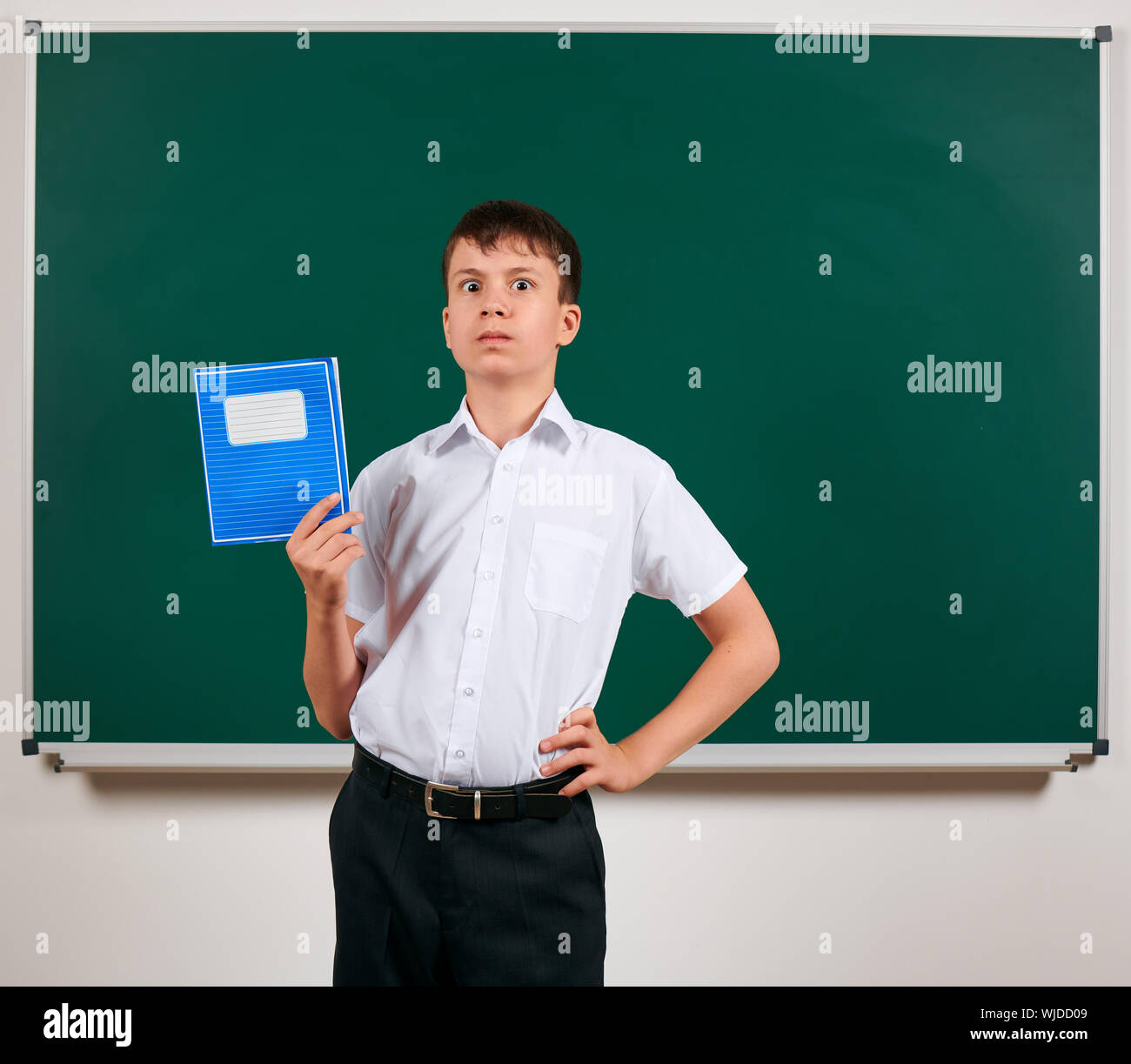 Portrait of a school boy posing with blue exercise book on blackboard ...