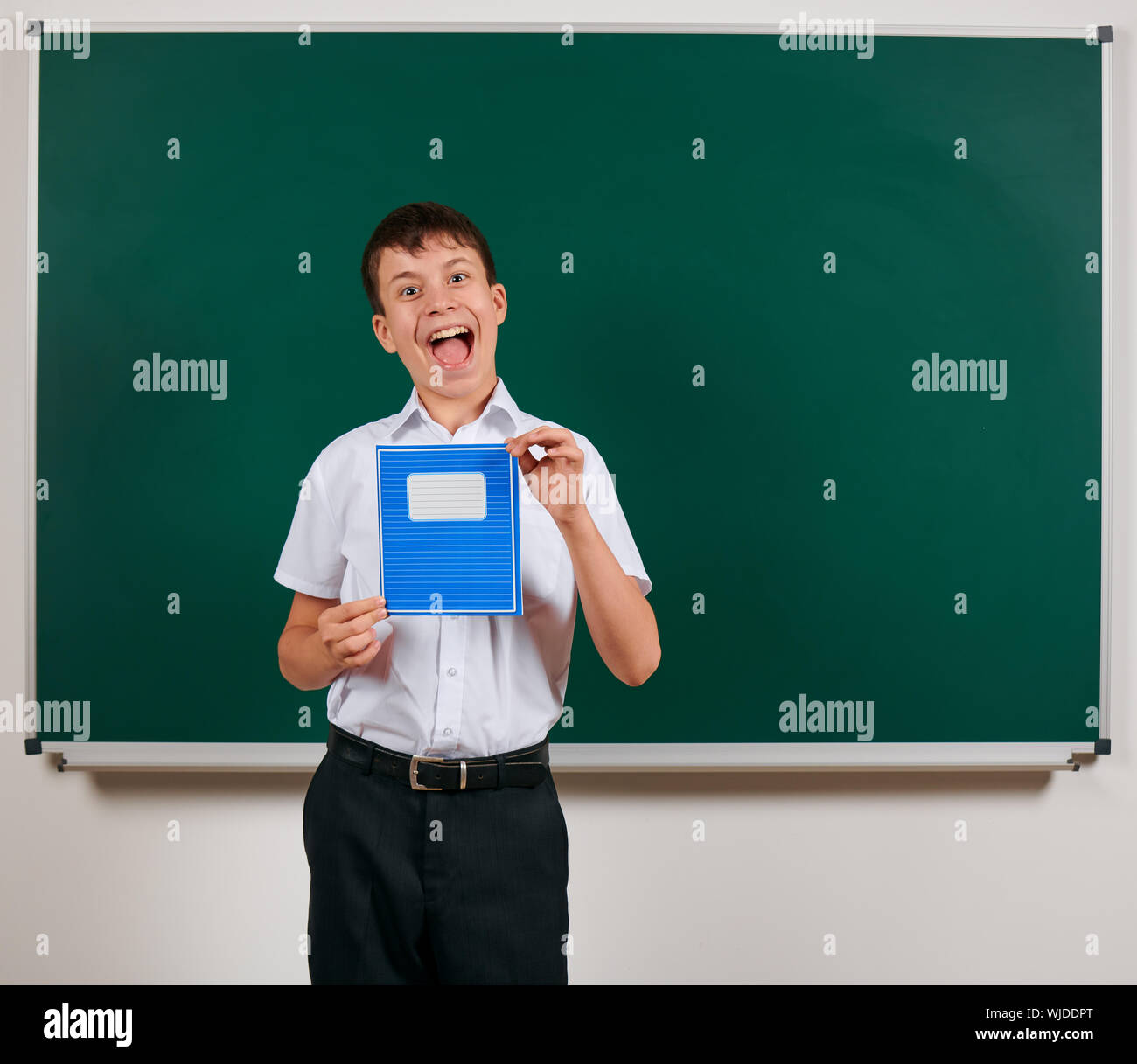 Portrait of a school boy posing with blue exercise book on blackboard ...