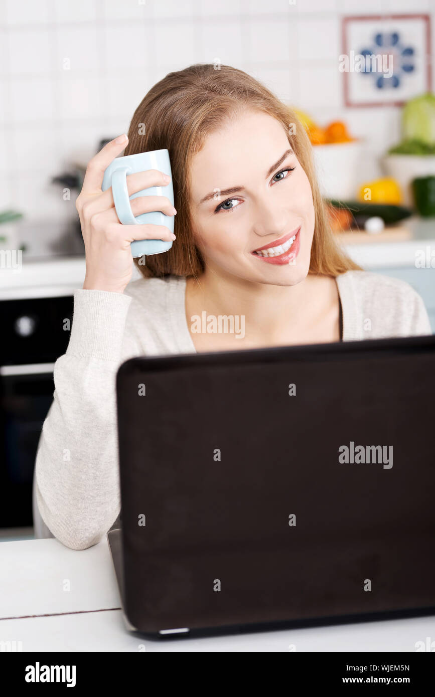 Young beautiful woman sitting by the table with laptop and cup. Kitchen ...