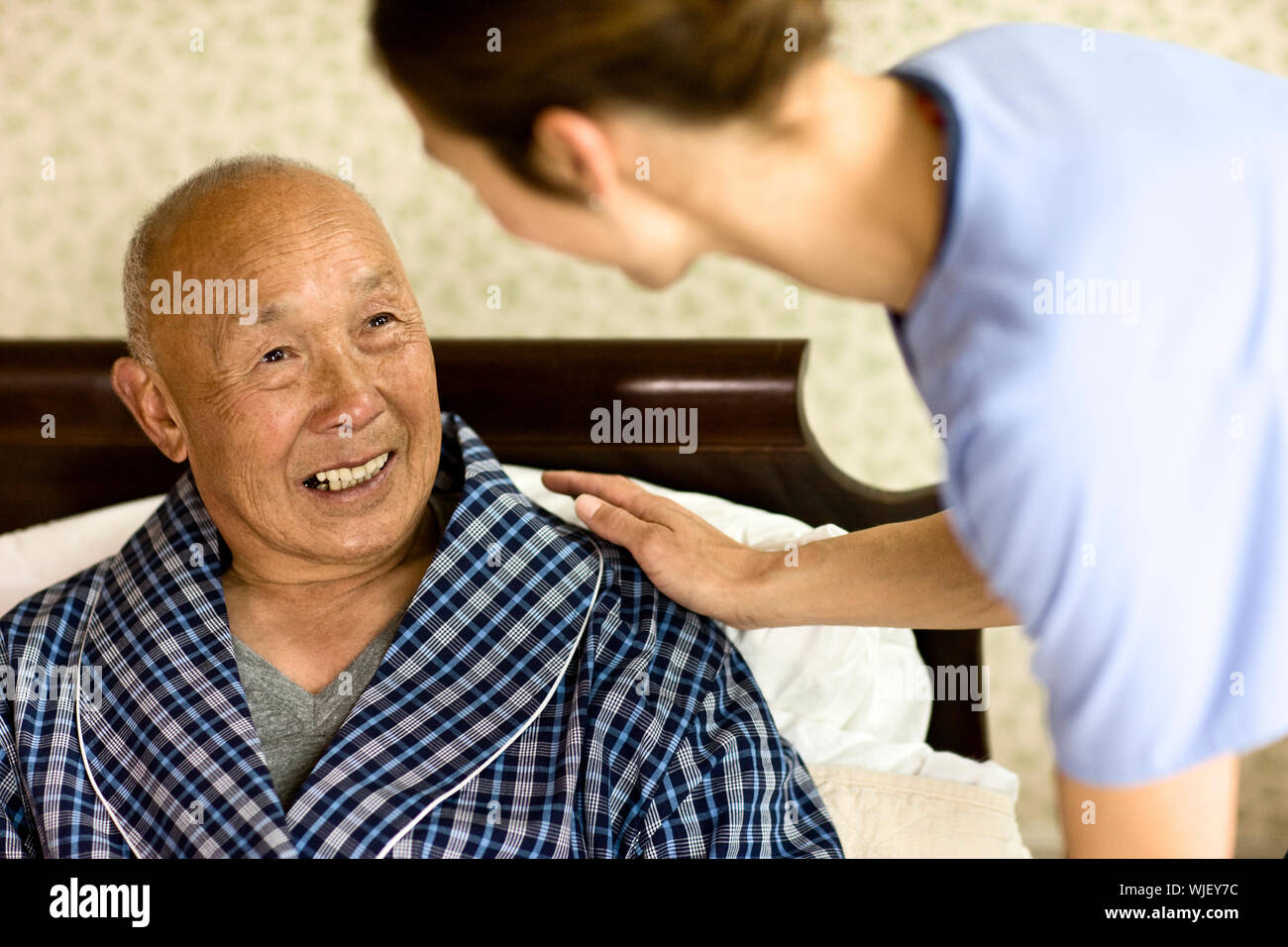 Senior man sitting in bed smiles up at a female nurse as she leans over ...