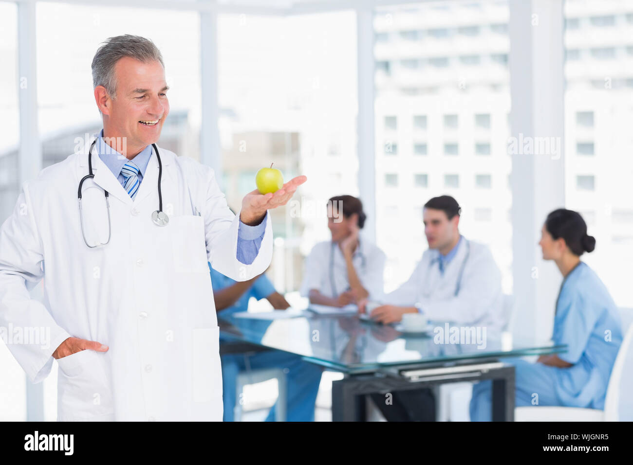 Portrait of a confident doctor holding apple with group around table in ...