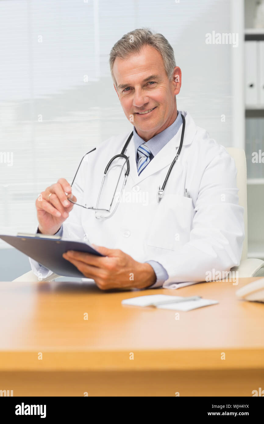 Smiling doctor sitting at his desk with clipboard his office at the ...