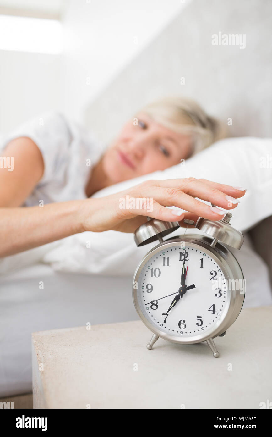 Blurred sleepy mature woman extending hand to alarm clock in bed at ...