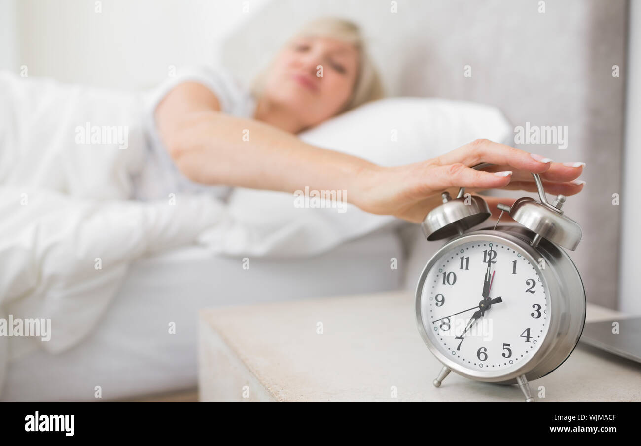 Blurred sleepy mature woman extending hand to alarm clock in bed at ...